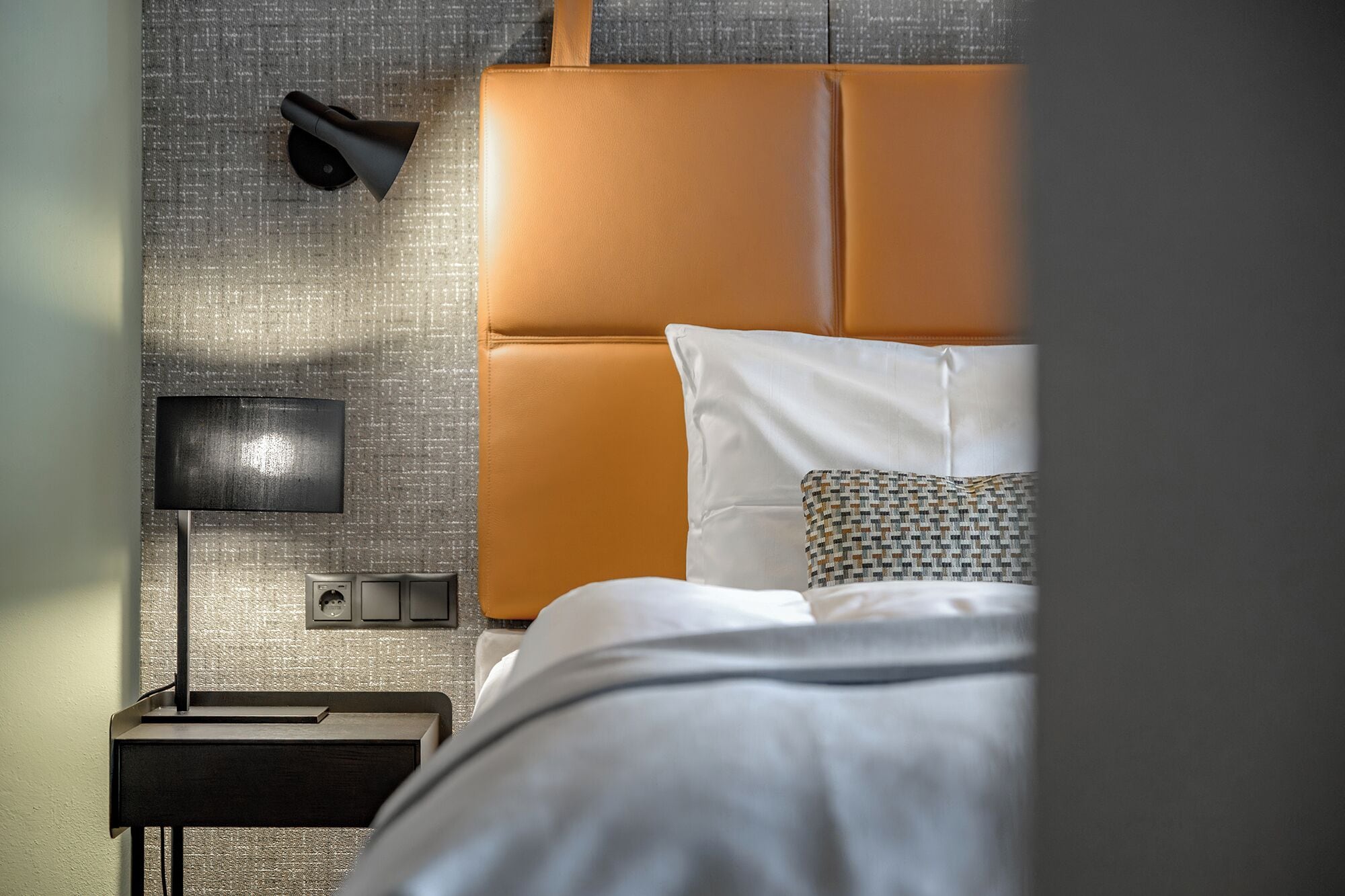 Quarto de hotel moderno com parede texturizada, cabeceira de couro, mesa de cabeceira com luminária e almofada estampada.