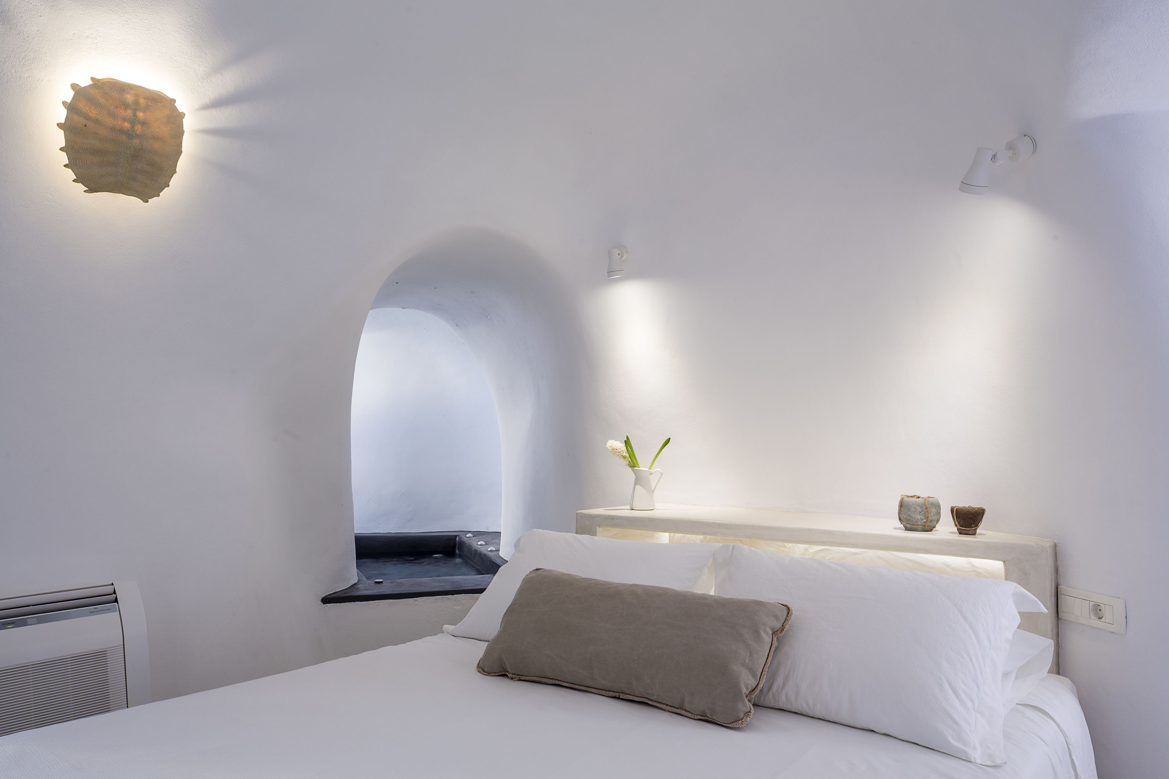 Quarto moderno com cama, mesa de cabeceira, luminária na parede e janela com vista em arco.