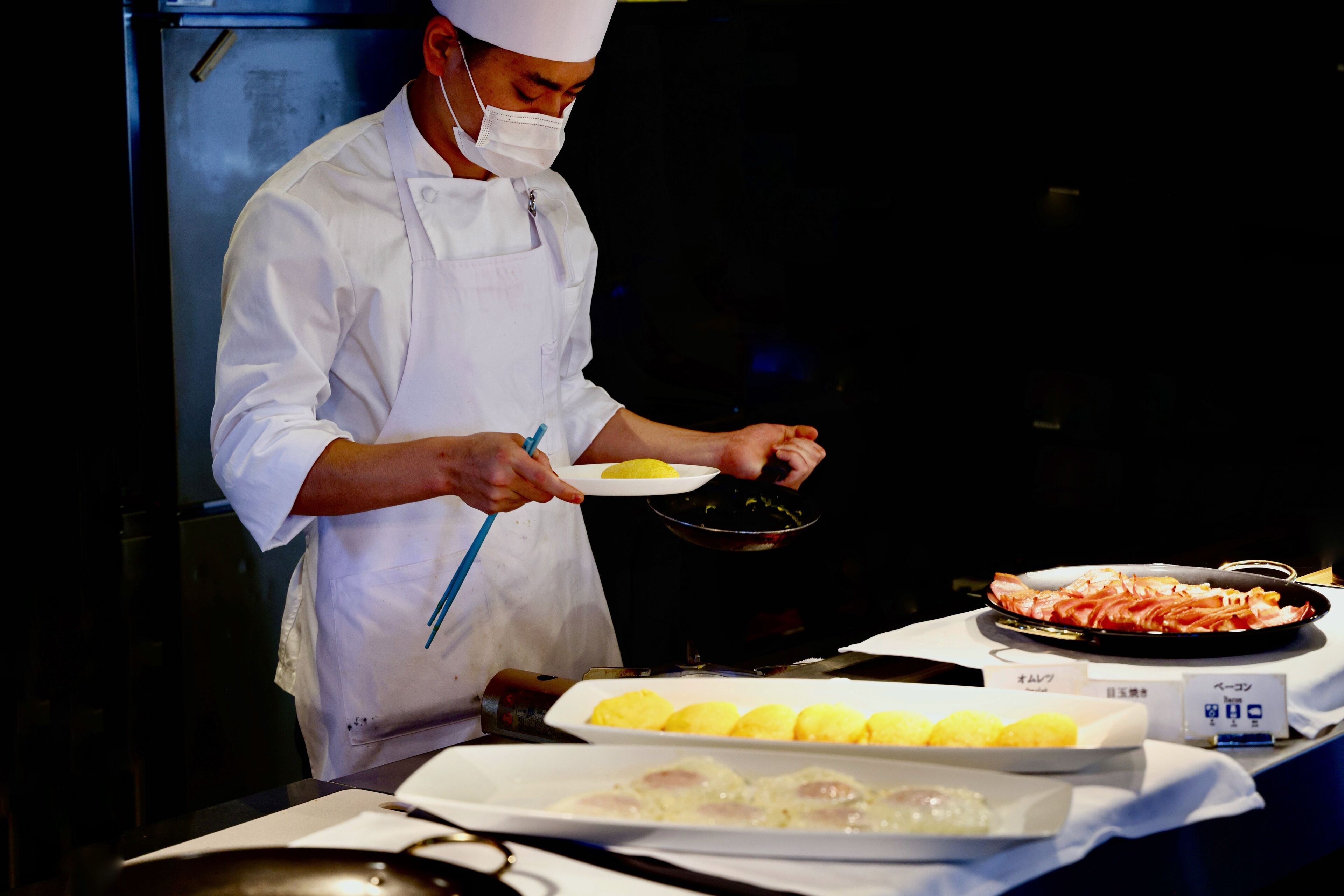 Um chef com uniforme branco prepara comida em uma mesa com diversos pratos, incluindo um prato com itens amarelos e uma bandeja de comida.