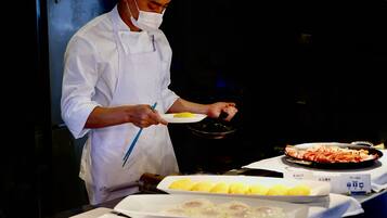 Um chef com uniforme branco prepara comida em uma mesa com diversos pratos, incluindo um prato com itens amarelos e uma bandeja de comida.