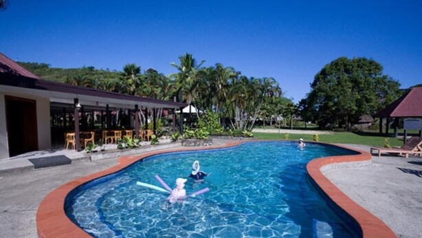 Piscina com borda curva, rodeada por um deck de concreto. Há cadeiras de descanso e uma área de estar coberta com mesas e cadeiras.