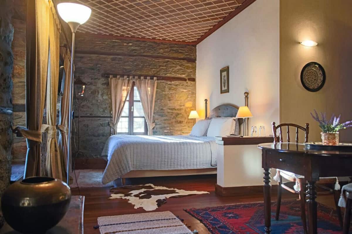 Quarto rústico com parede de pedra, uma cama com cabeceira, mesa de madeira e um abajur.