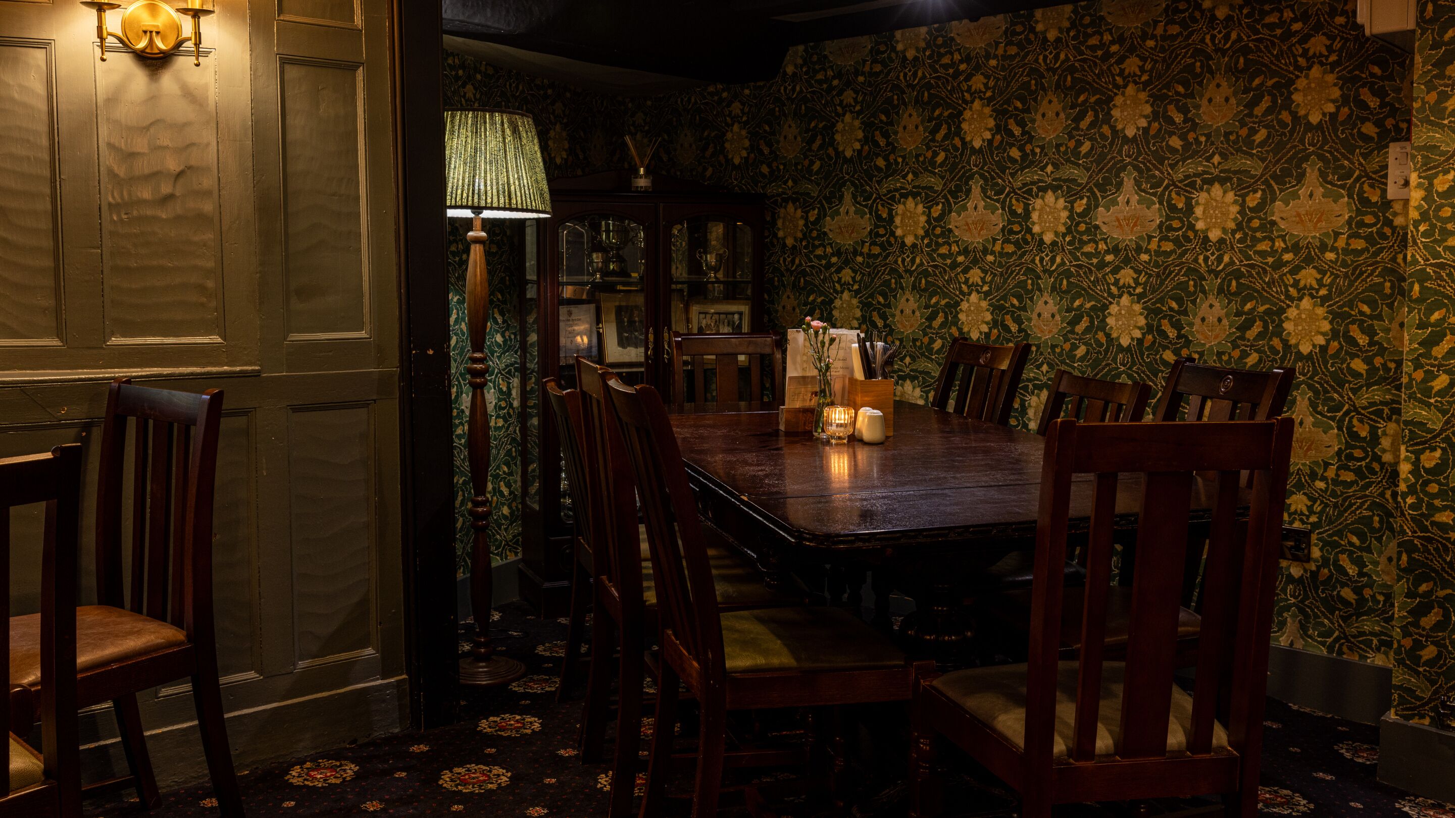 Uma sala de jantar com mesa de madeira, cadeiras, um abajur e papel de parede estampado.