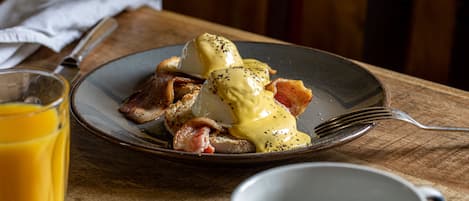 Prato de café da manhã com ovos Benedict, bacon e um copo de suco de laranja sobre uma mesa de madeira.