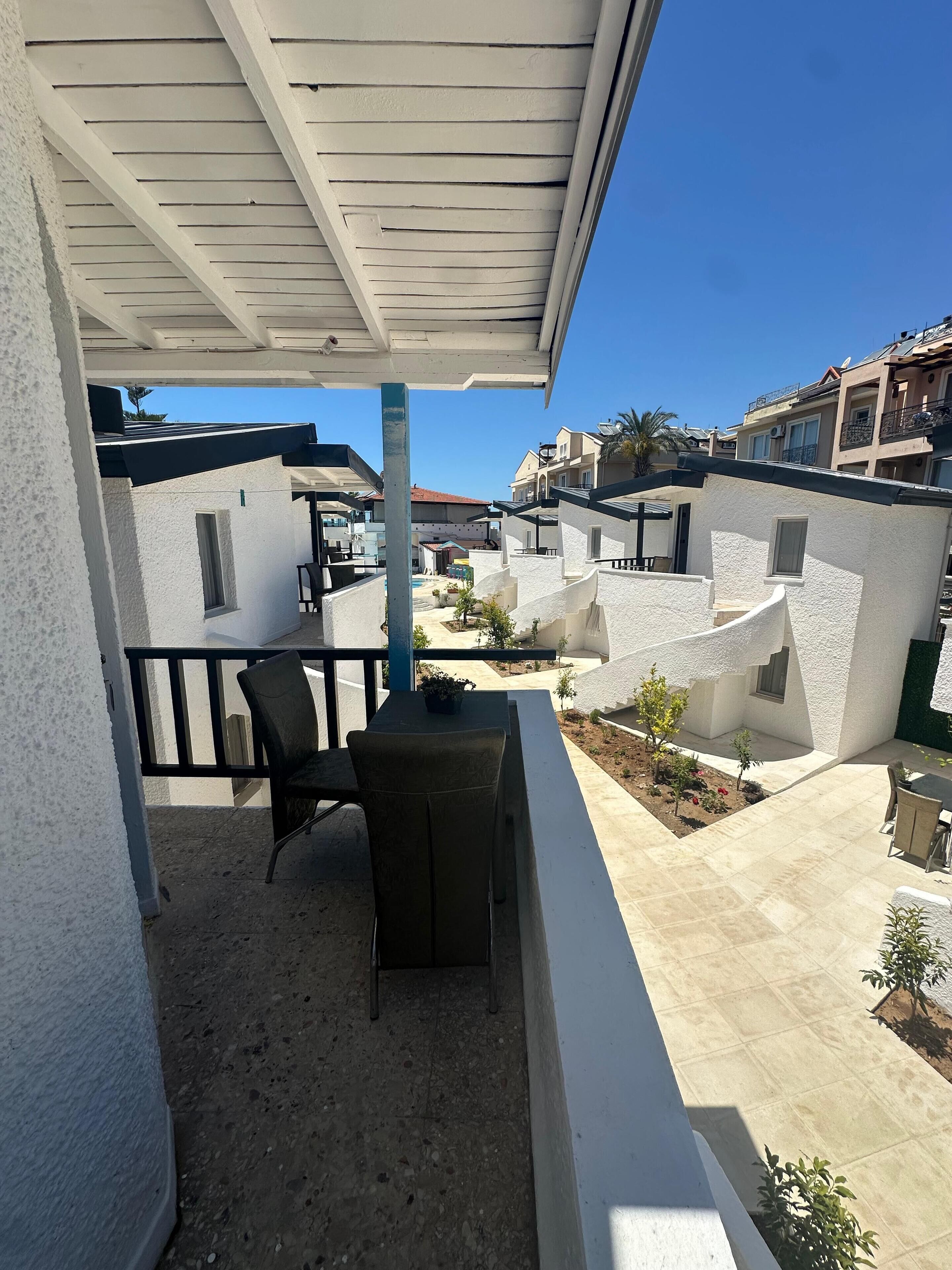 Uma área externa coberta com mesa e cadeiras, vista para casas vizinhas e um céu azul claro.