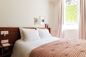 Cama bem arrumada com lençóis brancos e manta xadrez, mesa de cabeceira com tomada e duas fotografias emolduradas na parede.