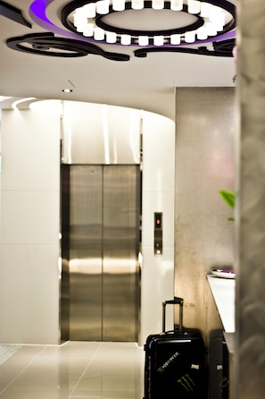 Um elevador com acabamento metálico e um painel de controlo na parede. Uma mala preta com logótipo verde está colocada perto do elevador.
