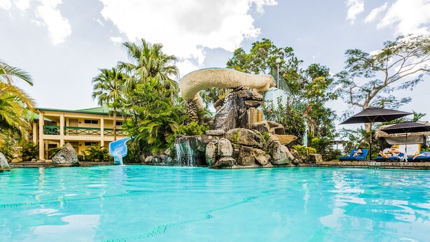 Piscina com cachoeira artificial de pedra, rodeada por palmeiras e um edifício com telhado verde.