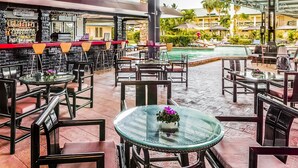 Uma área de bar ao ar livre com várias mesas e cadeiras, piscina ao fundo e paisagem tropical com palmeiras.