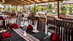 Um restaurante ao ar livre com mesas e cadeiras de madeira, uma piscina ao fundo e um jardim tropical.