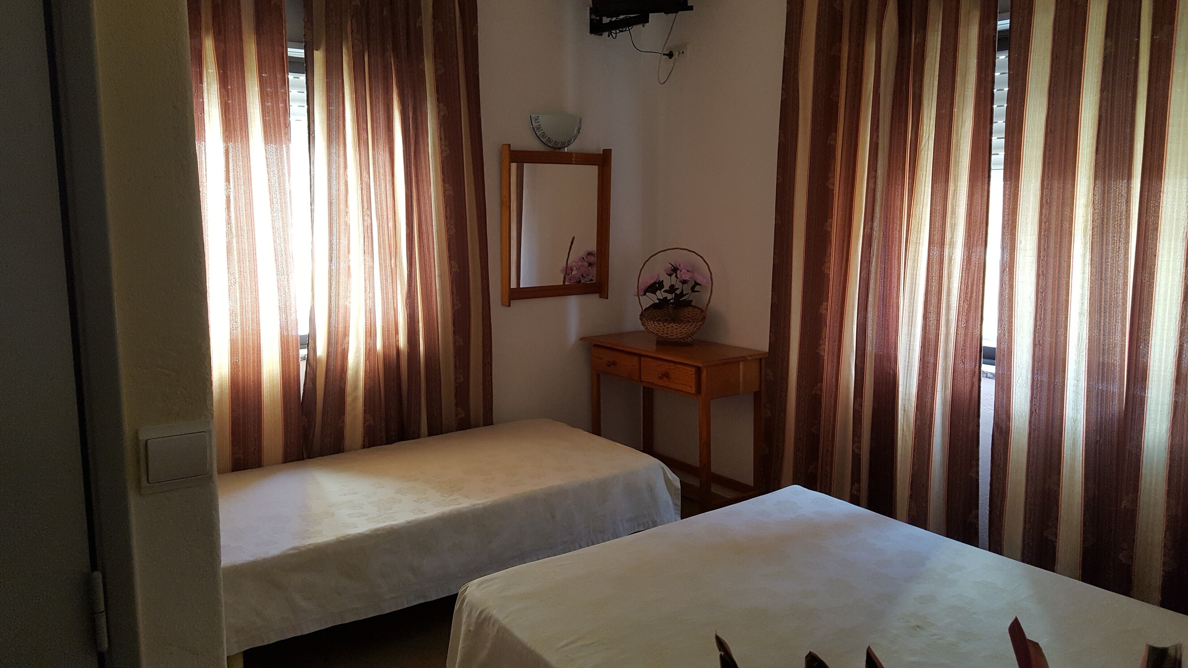 Quarto com duas camas, uma mesa de cabeceira de madeira com um cesto de flores, um espelho fixado na parede e cortinas listradas.