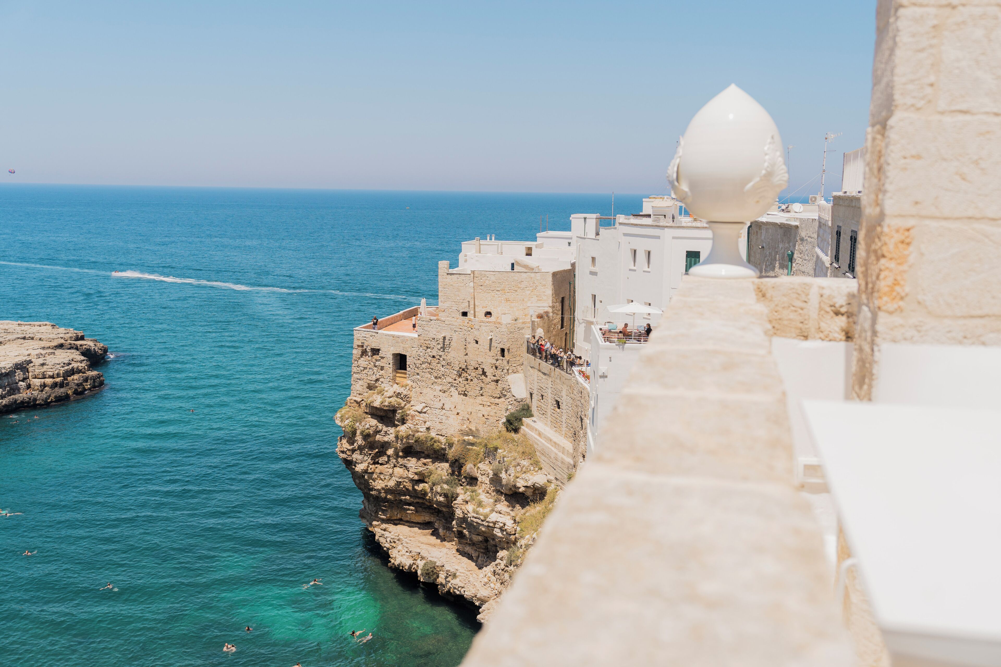 Uma vila costeira com edifícios de pedra, mar azul cristalino e uma estrutura esférica branca imponente.