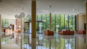 Um hall amplo com balcão de recepção, área de estar e grandes janelas com vista para o verde.