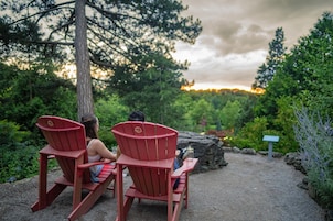 Duas pessoas sentadas em cadeiras Adirondack vermelhas, olhando para uma paisagem natural com um caminho de cascalho e um sinalizador.