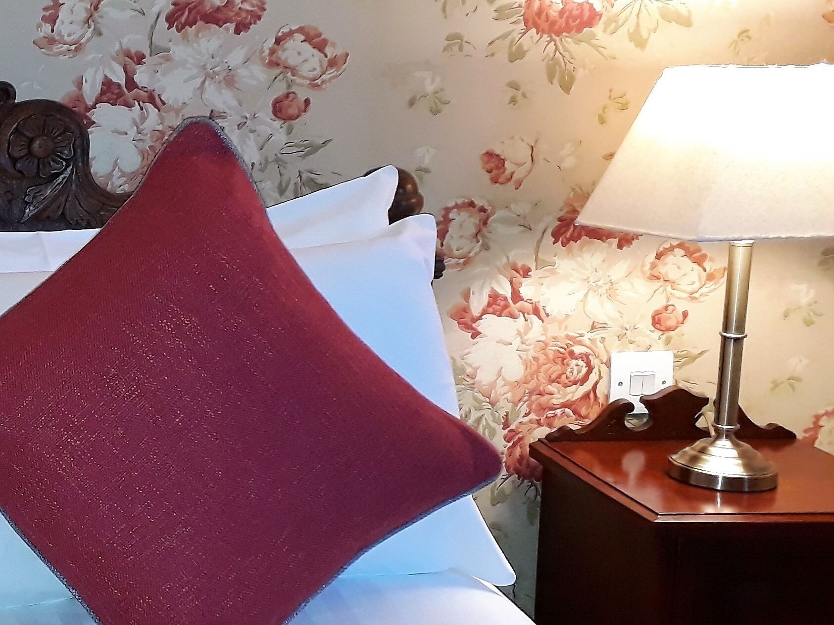 Uma cama com travesseiro vermelho, lençóis brancos e papel de parede floral.