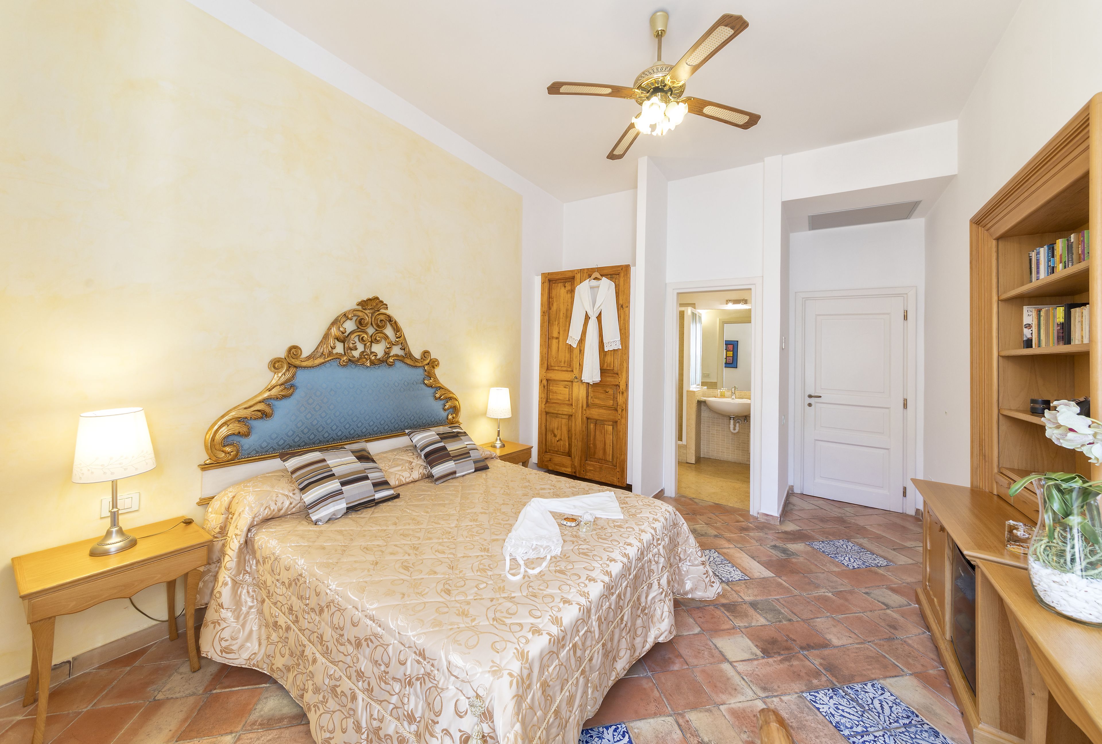 Quarto de hotel com cama grande, cabeceira de madeira, ventilador de teto, mesa de cabeceira com abajur, guarda-roupa de madeira e estante.