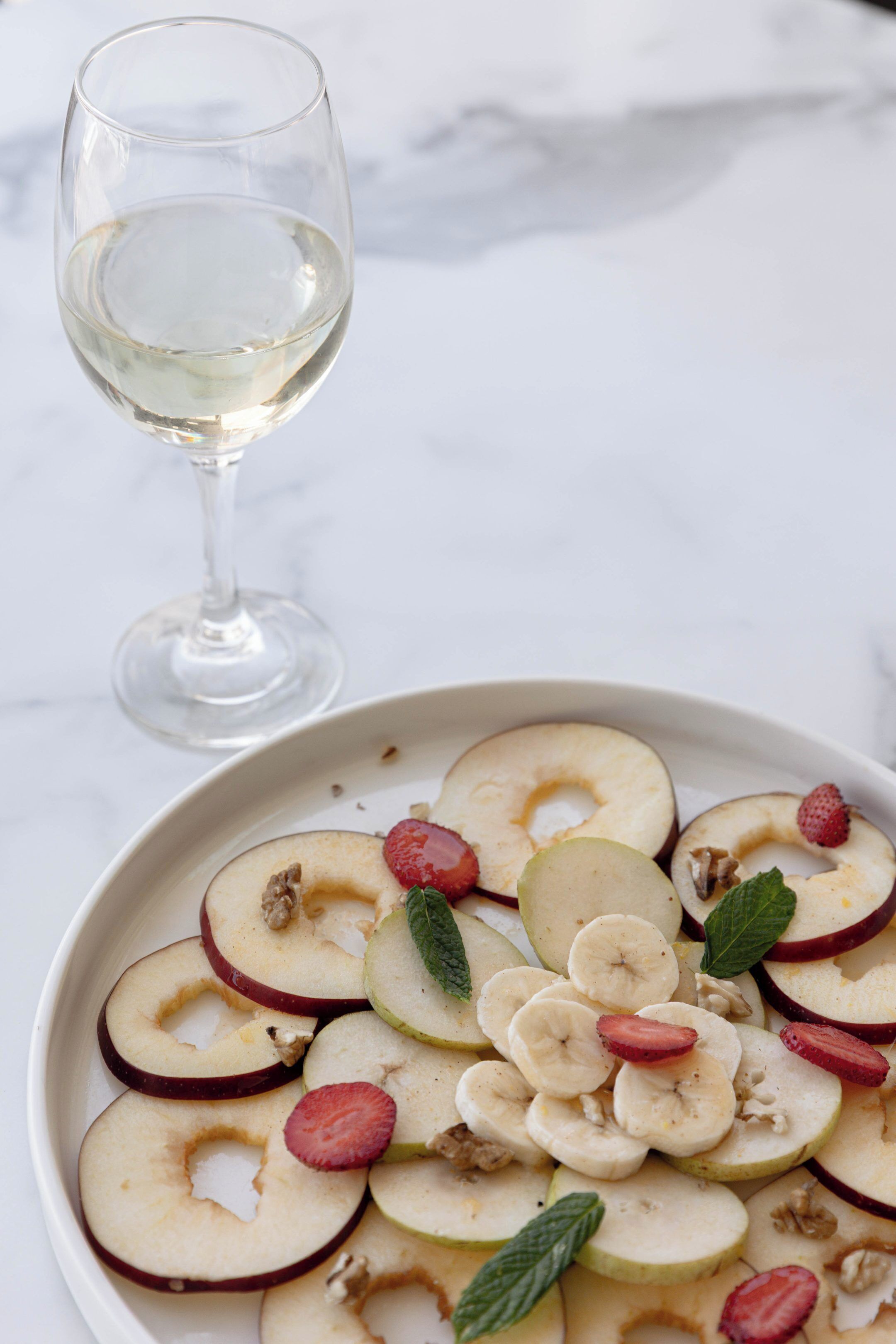 Um prato de fatias de maçã, banana e morangos, com nozes e folhas de hortelã, acompanhado de um copo de vinho branco.