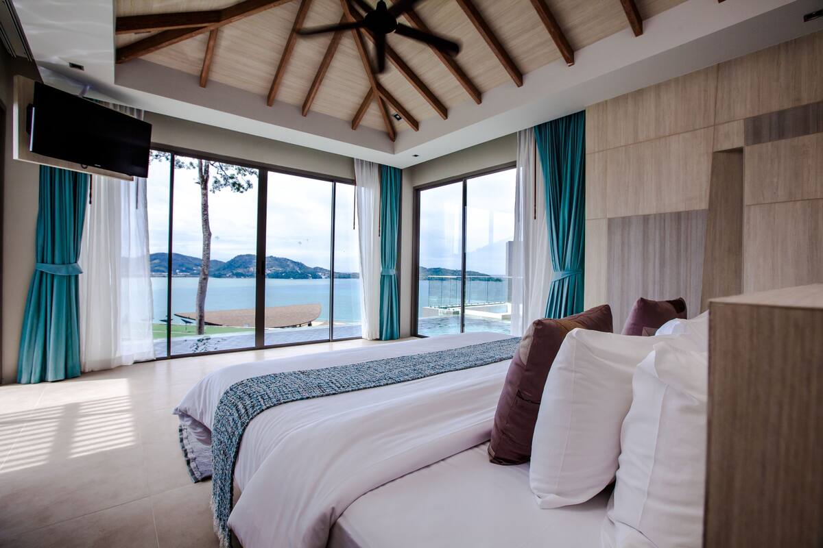 Quarto com uma cama grande, uma televisão na parede e vista para o mar através de amplas janelas.