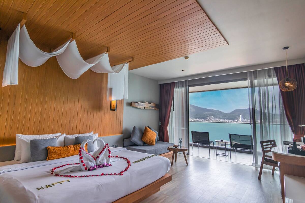Quarto de hotel com cama grande, sofá, varanda vista para o mar e área de refeições.
