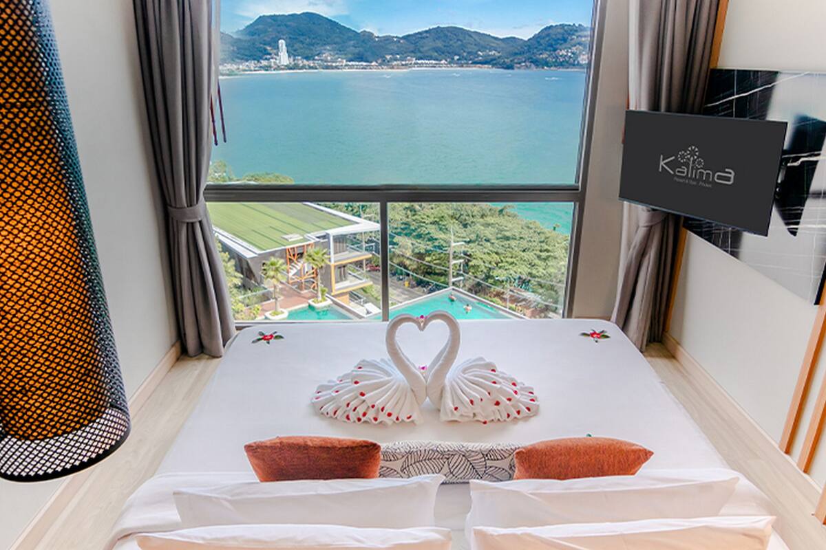 Quarto de hotel com cama adornada com decoração em formato de coração, vista para um corpo de água e montanhas, e uma placa com o nome do hotel.