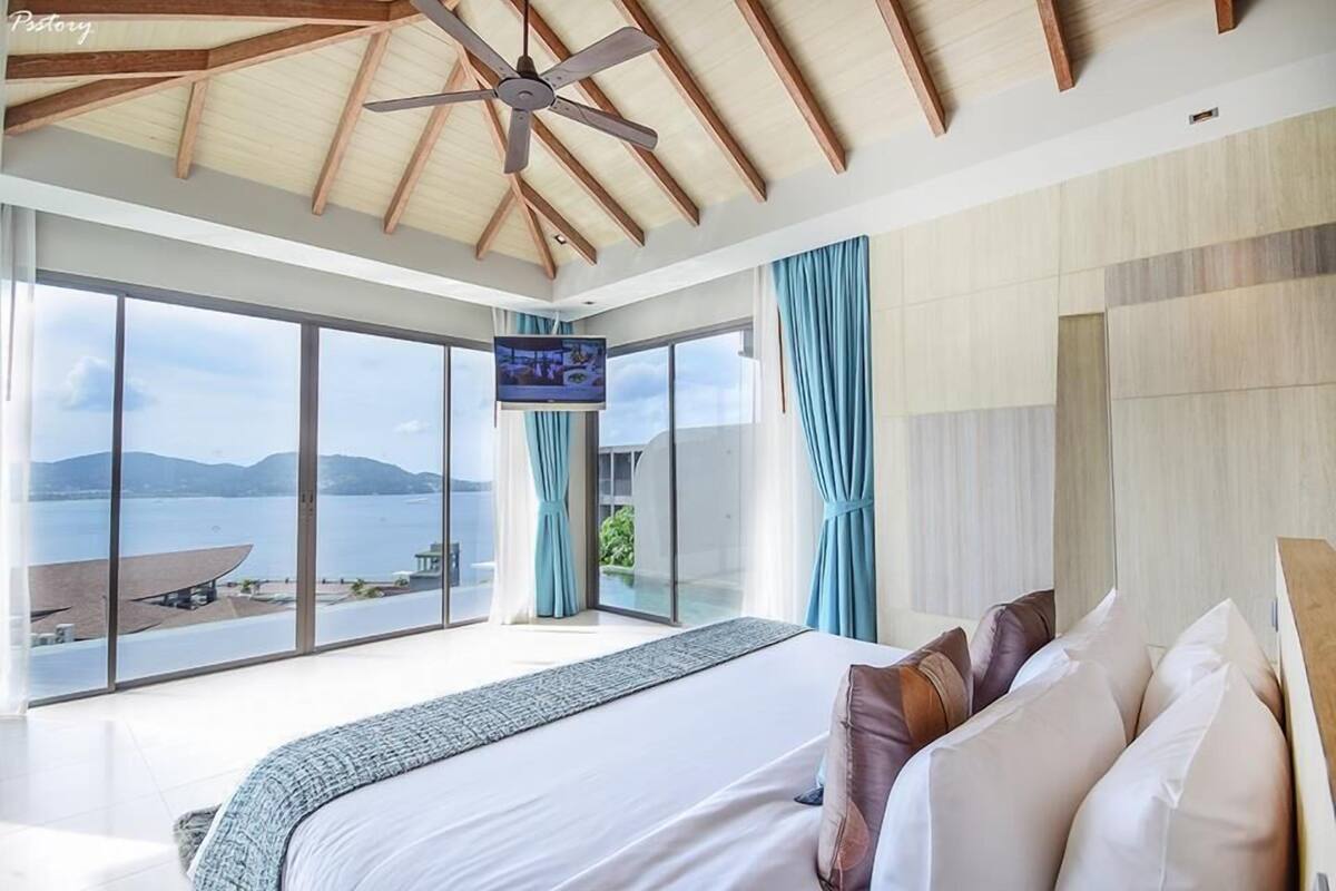 Quarto moderno com uma cama grande, ventoinha no teto e televisão. Possui uma janela ampla com vista para o mar e montanhas.