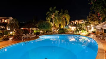 Piscina à noite com espreguiçadeiras e guarda-sóis, cercada por árvores e edifícios.