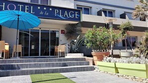 Entrada de hotel com guarda-sol azul, mesas ao ar livre e plantas em vasos.