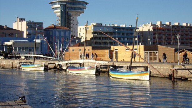 Uma orla com vários barcos atracados, uma torre e edifícios ao fundo.