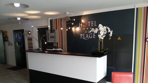 Uma receção de hotel com balcão em preto e branco, uma cadeira vermelha e uma parede com o nome do hotel e uma planta decorativa.
