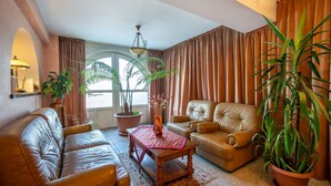 Sala de estar com sofá de couro, mesa de centro de madeira e poltrona de couro. Há plantas em vasos e uma janela grande com cortinas.