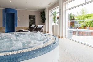 Uma banheira de hidromassagem com borda revestida, uma área de estar com duas cadeiras e uma janela grande com vista para uma piscina e vegetação.