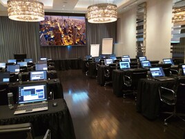 Sala de conferência com várias mesas, laptops e uma tela grande exibindo uma paisagem urbana.