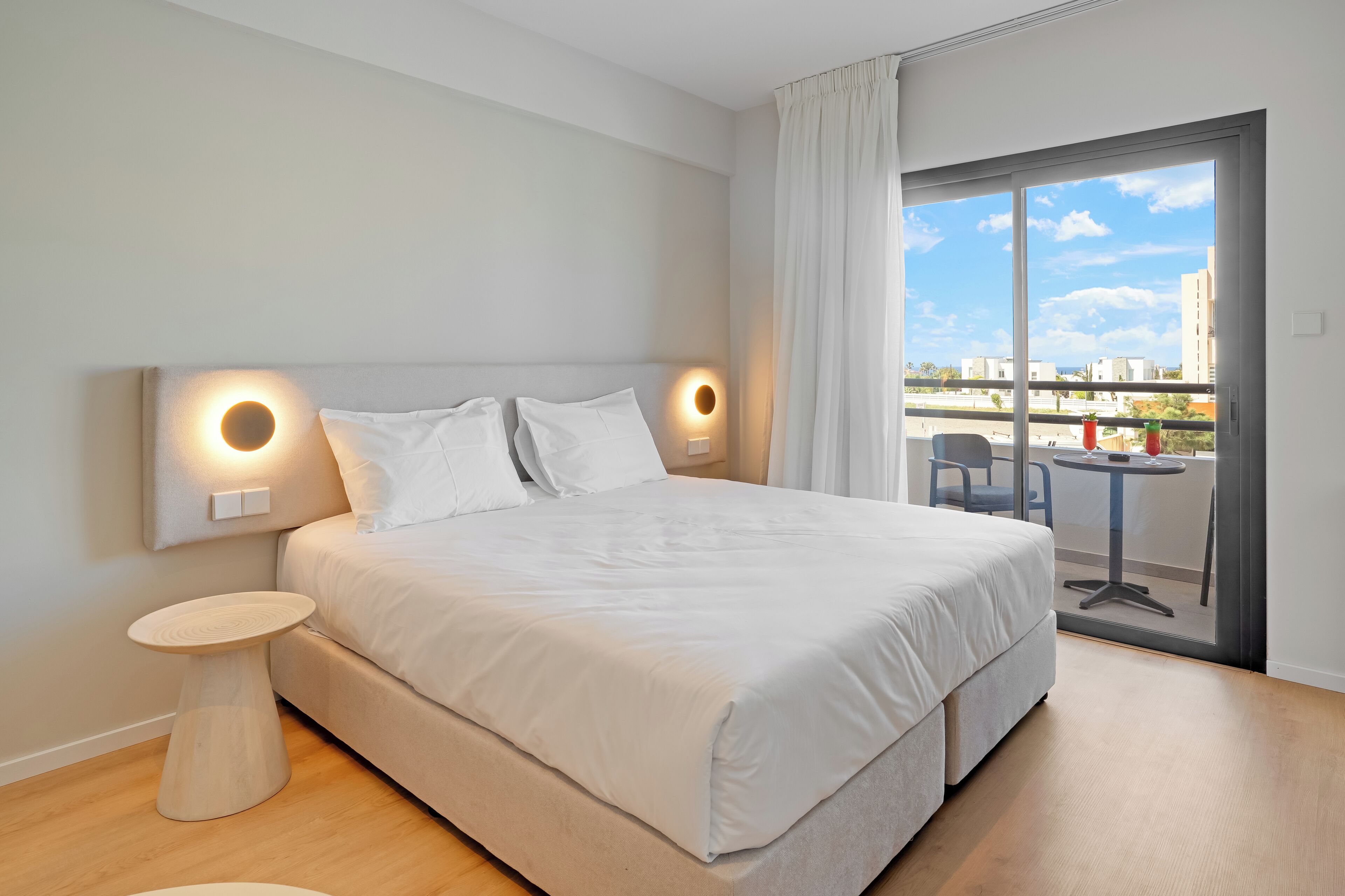 Quarto de hotel moderno com uma cama grande, duas mesas de cabeceira, uma mesa redonda pequena e uma varanda com vista.