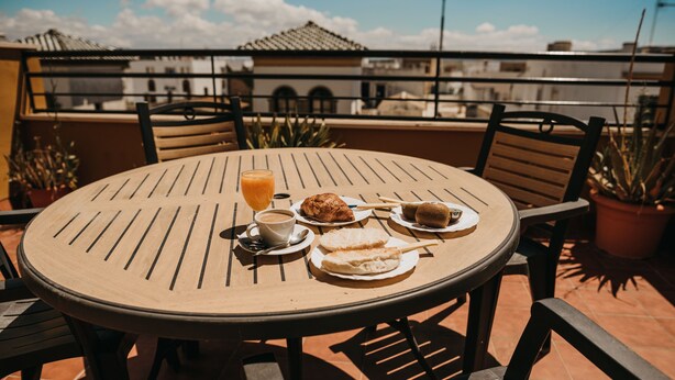Mesa redonda com bebida, doces e sanduíche num terraço com cadeiras e plantas em vasos.