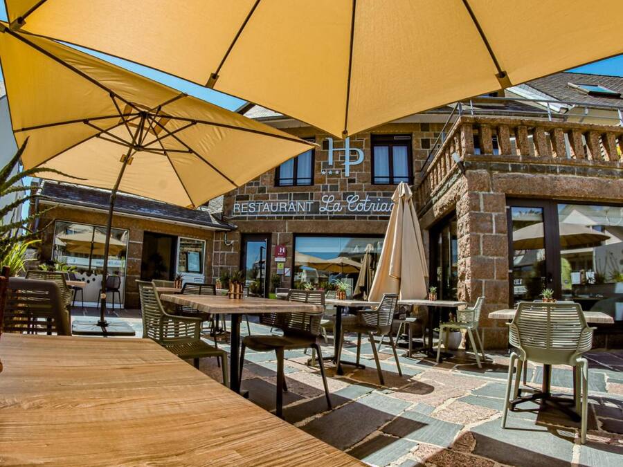 Uma área externa com mesas e cadeiras sob grandes guarda-sóis, um edifício de pedra com placa de restaurante e um deck de madeira.
