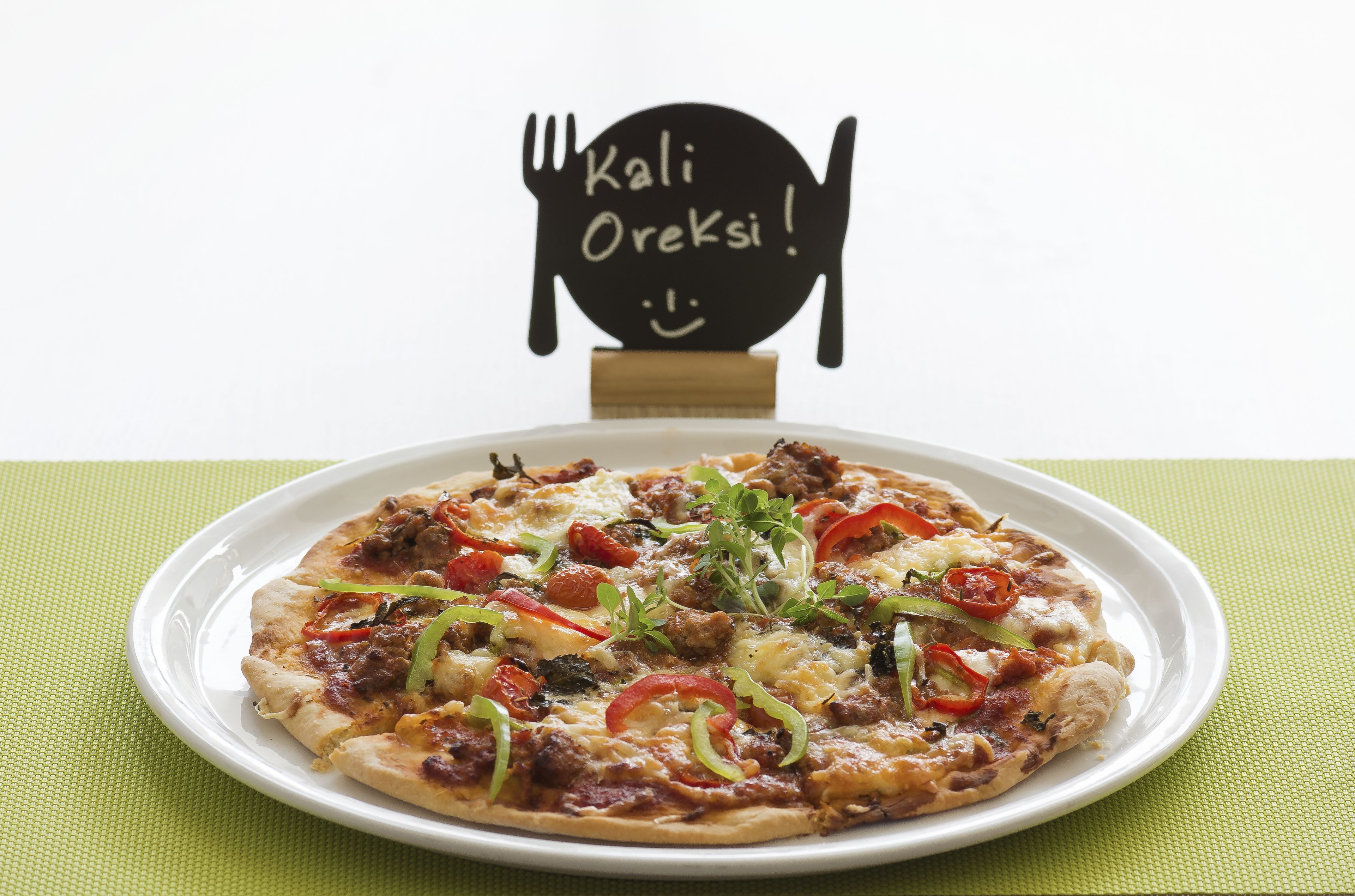 Pizza em um prato branco com cobertura de pimentões verdes e tomates, sobre uma toalha de mesa verde.
