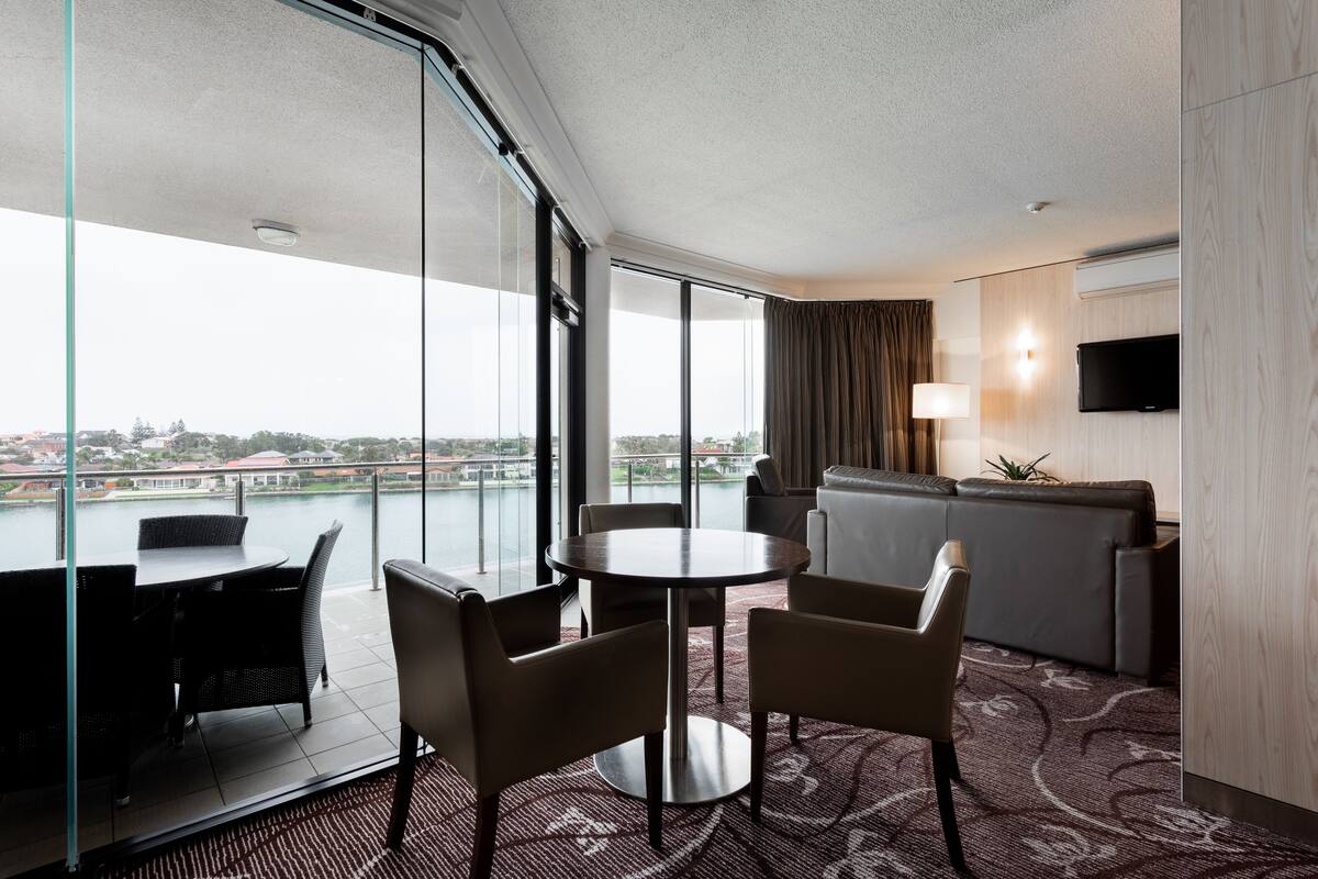 Quarto de hotel com varanda, uma mesa redonda, quatro cadeiras, um sofá, uma TV e vista para a água e edifícios.