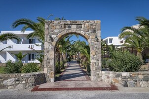 Um arco de pedra que dá acesso a um caminho flanqueado por palmeiras e edifícios.