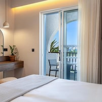 Quarto de hotel com cama, escrivaninha com luminária, planta em vaso e porta de vidro deslizante para área externa com cadeira e mesa.