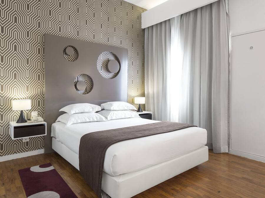 Quarto moderno com papel de parede estampado, uma cama grande com roupa de cama branca, duas mesas de cabeceira com abajures e piso de madeira.
