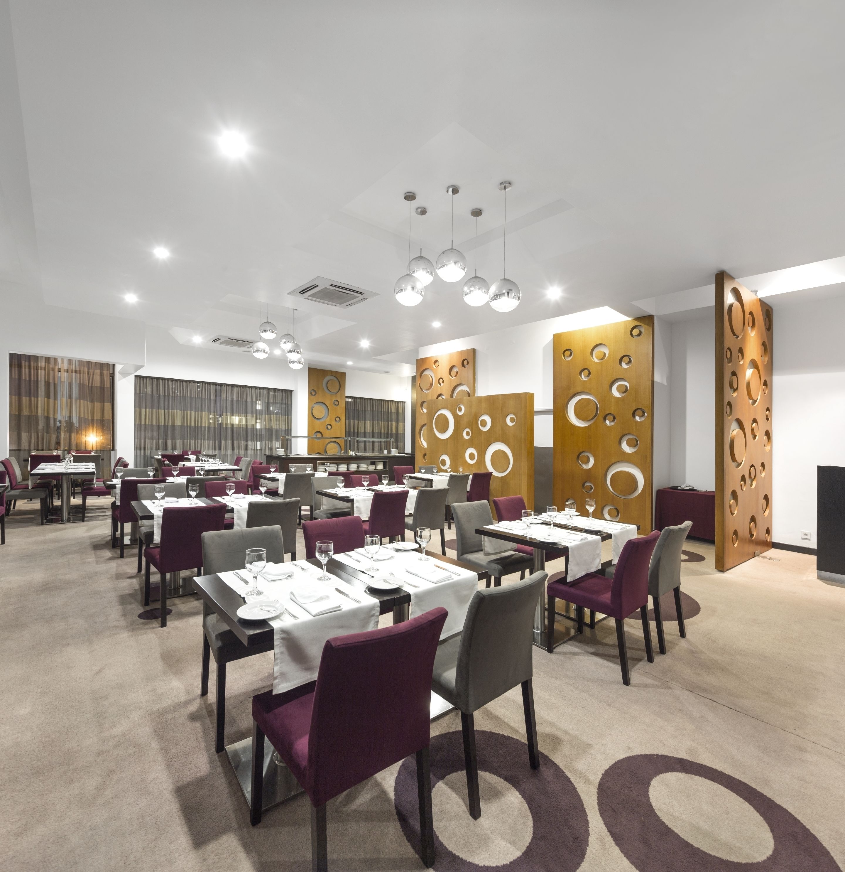 Espaço de refeições de hotel com várias mesas postas, iluminação pendente moderna e painéis decorativos na parede.