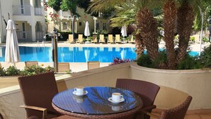 Uma área à beira da piscina com cadeiras de vime, uma mesa de vidro e vista para a piscina e edifícios do hotel.