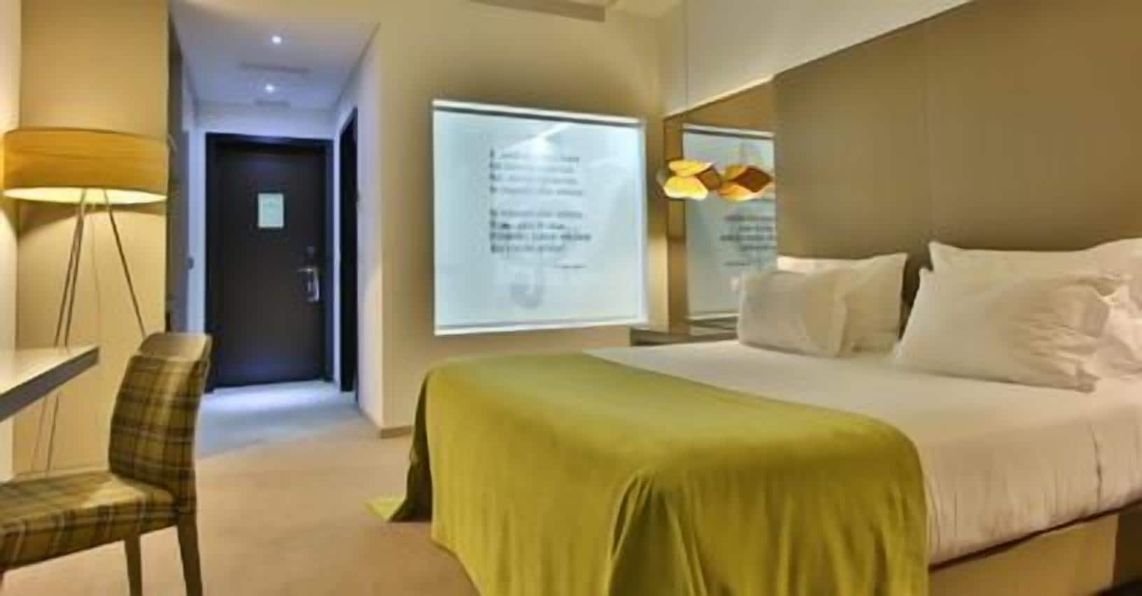 Quarto de hotel com cama, escrivaninha, cadeira e tela na parede exibindo texto.
