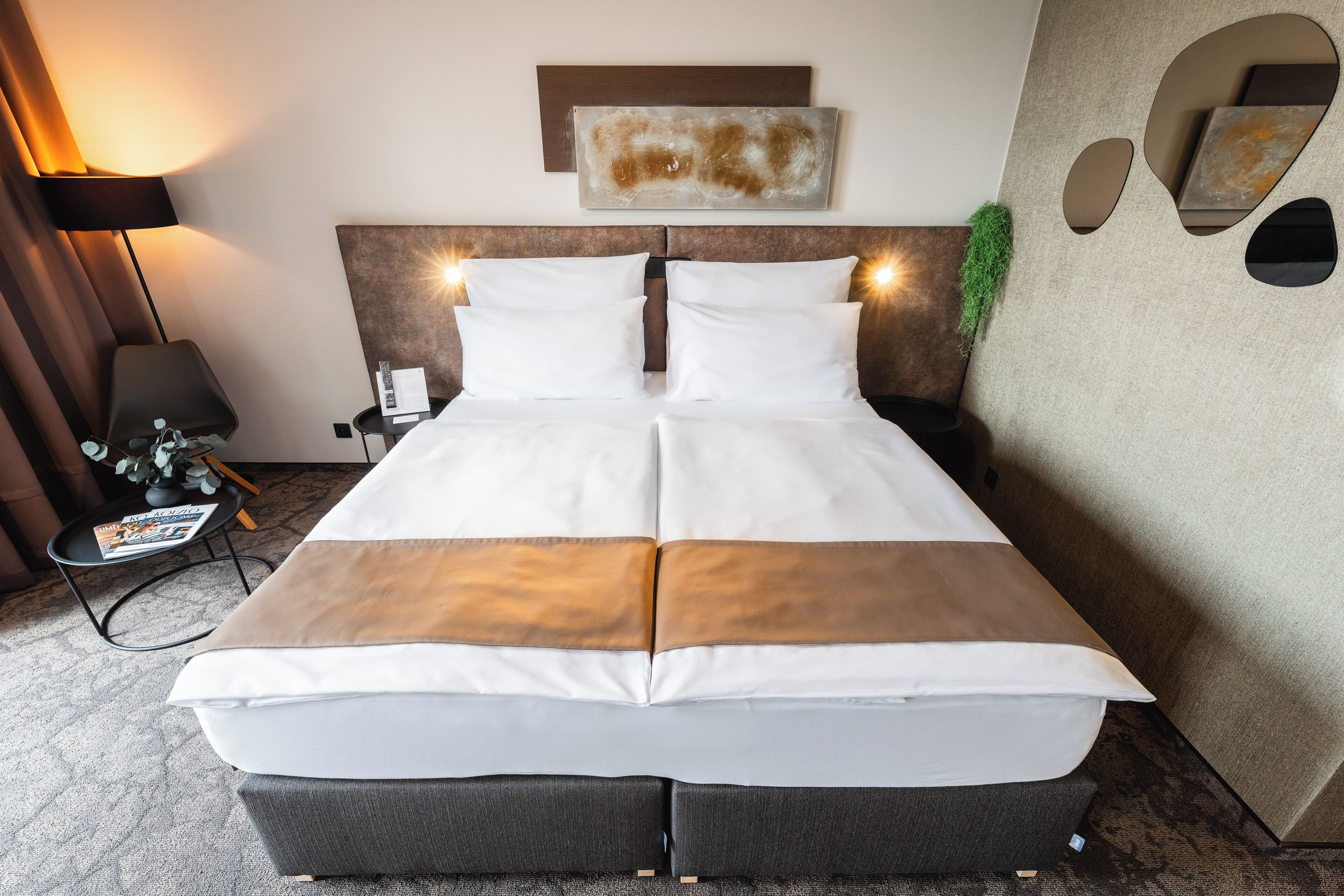 Uma cama de casal com cabeceira, mesa de cabeceira com abajur e uma revista sobre ela.