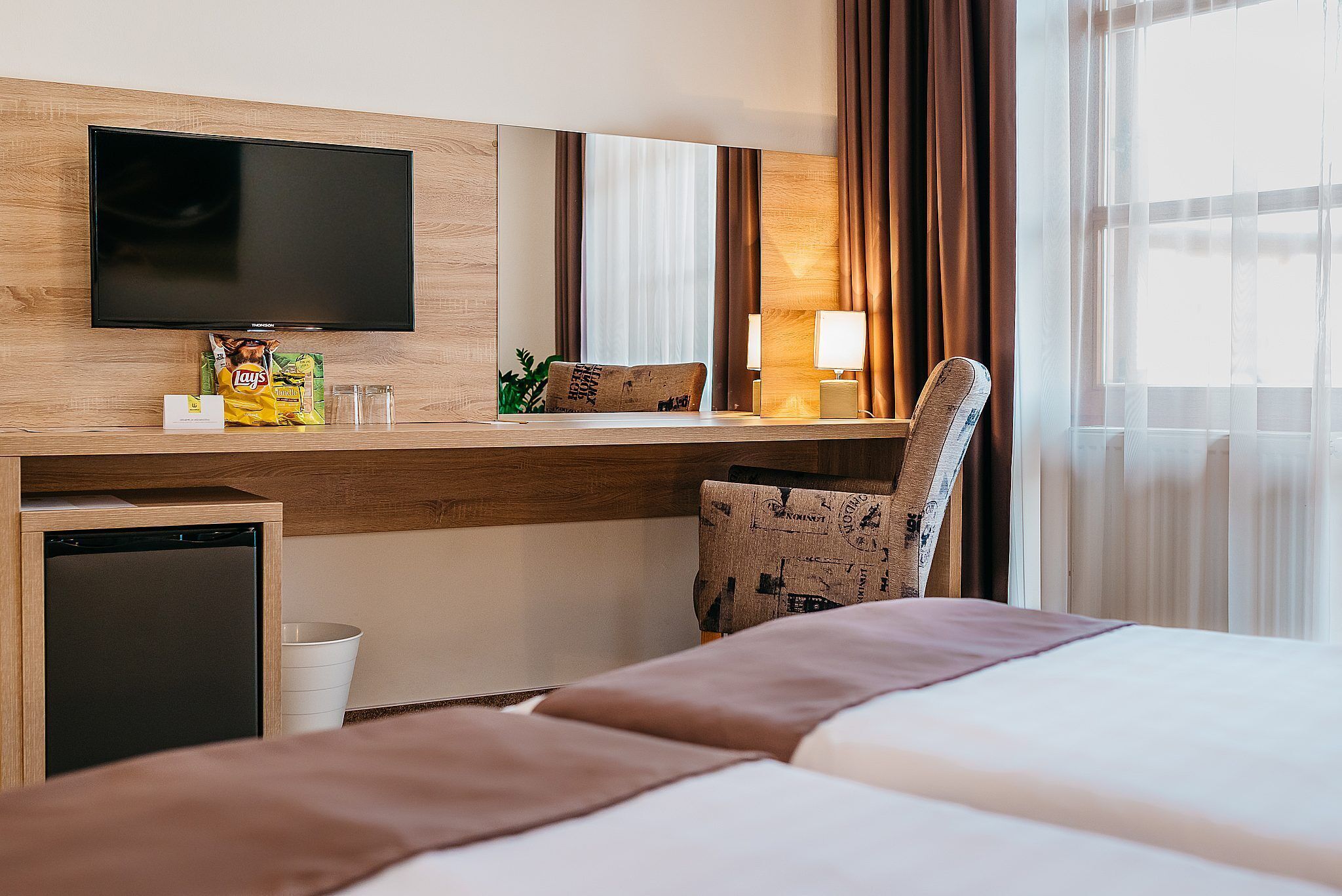 Quarto de hotel com cama, secretária com televisão de tela plana, cadeira, abajur e janela com cortinas.