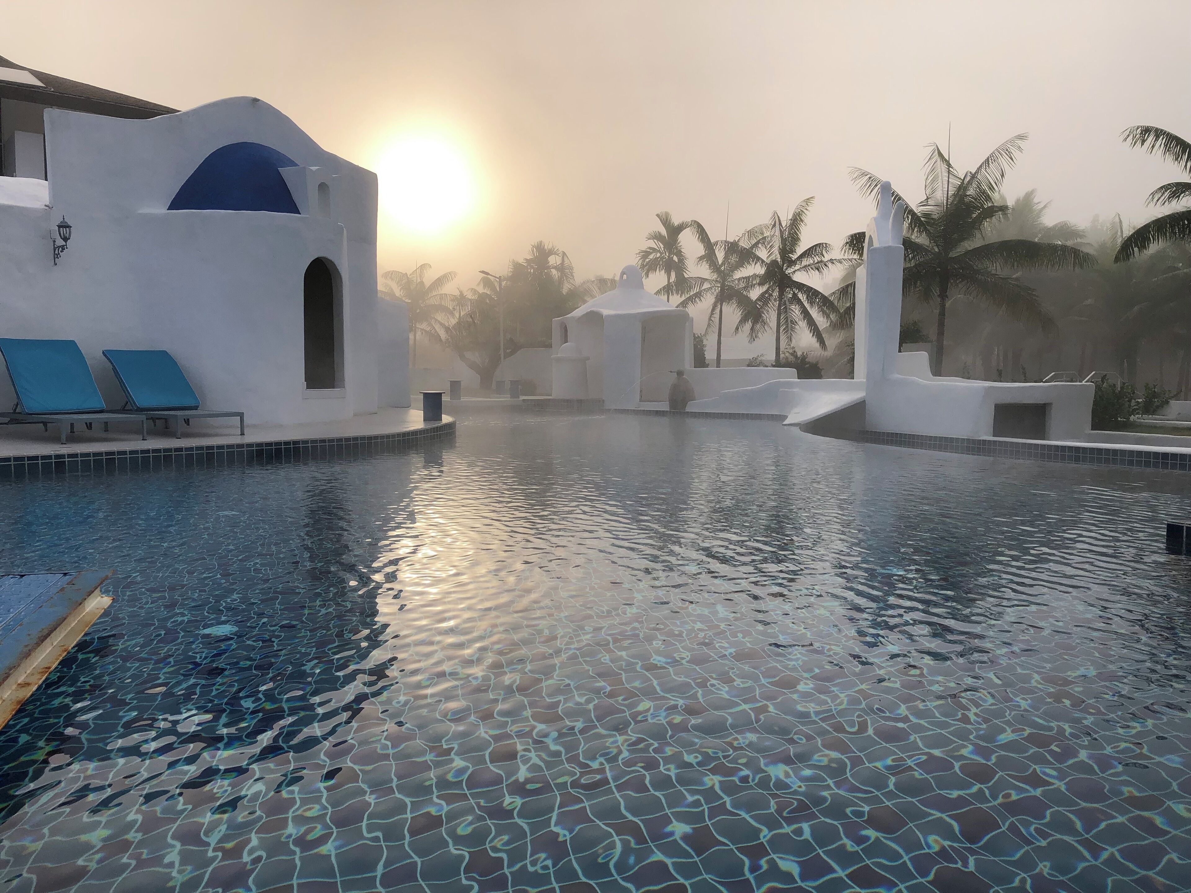 Piscina com água cristalina, rodeada por edifícios brancos e palmeiras.