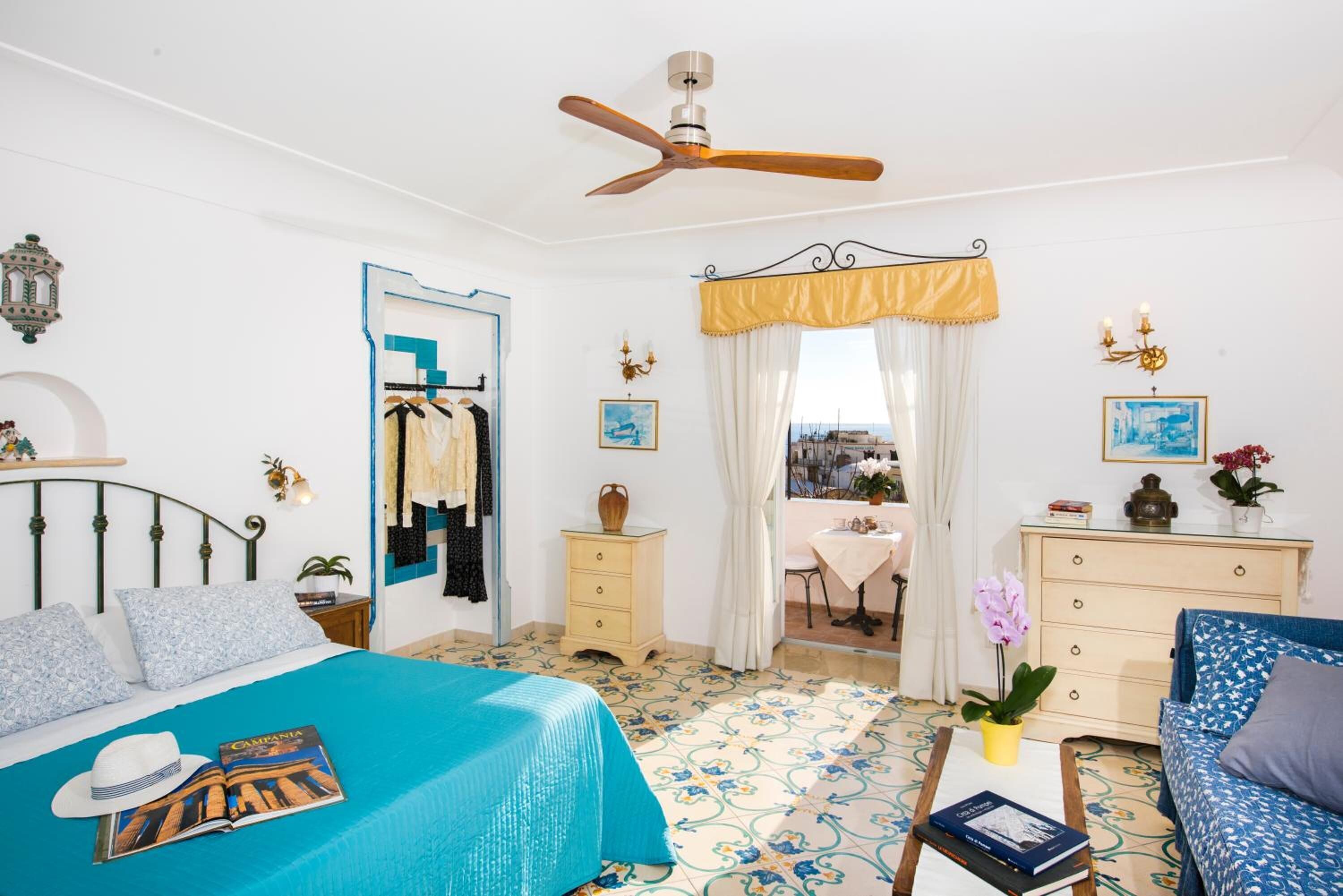 Um quarto com cama, ventilador de teto, janela com cortinas, uma mesinha e um sofá.