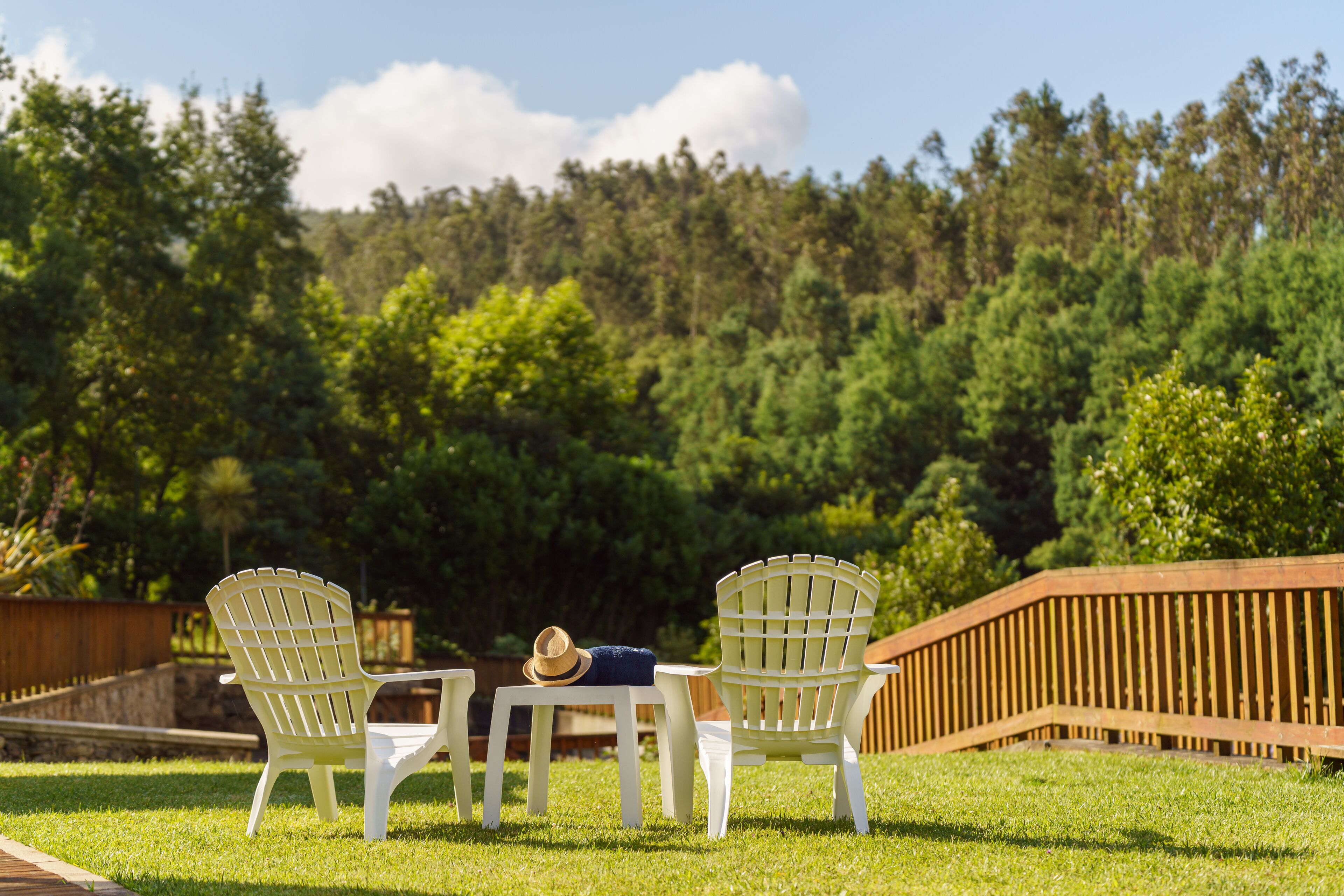 Duas cadeiras Adirondack, uma mesinha e um chapéu sobre a mesa, numa área externa gramada com uma cerca de madeira e uma colina arborizada ao fundo.