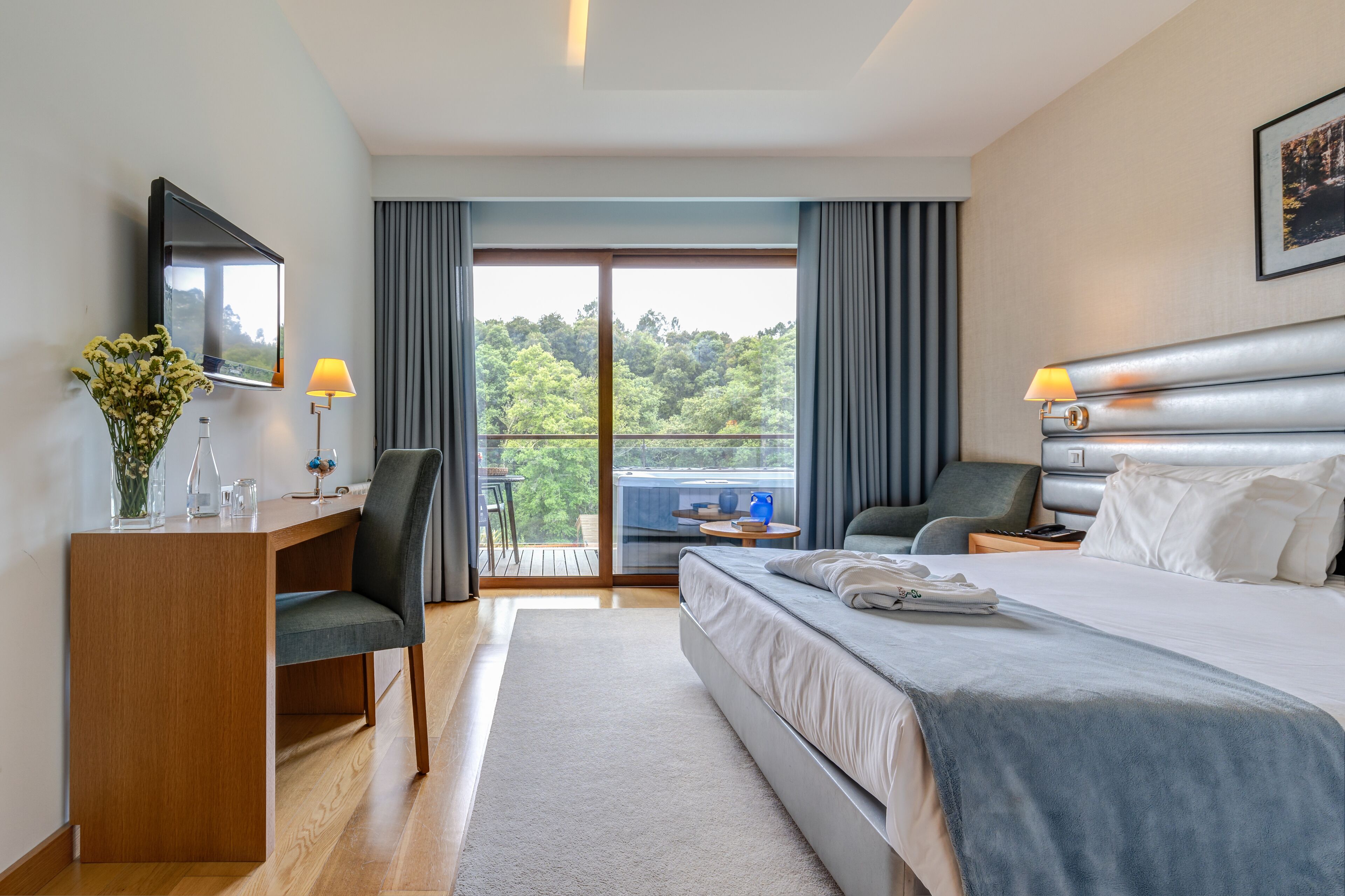 Quarto de hotel moderno com uma cama grande, uma escrivaninha, uma cadeira, uma televisão e uma varanda com vista para árvores.