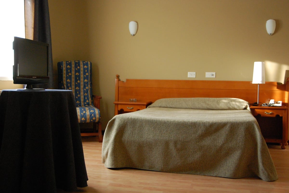 Quarto de hotel com uma cama, televisão sobre uma mesa, uma cadeira e mesa de cabeceira com abajur.